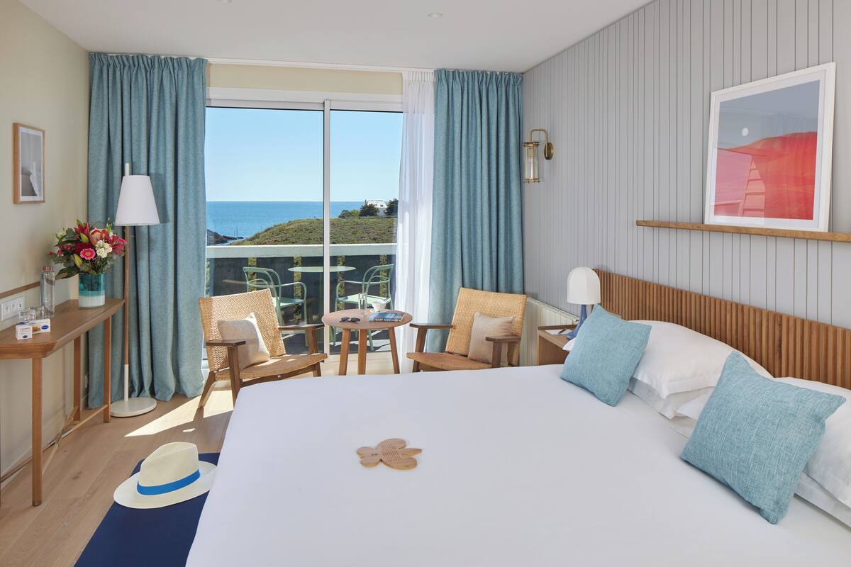 Quarto de hotel com uma cama grande, duas cadeiras, uma mesinha e vista para o oceano.