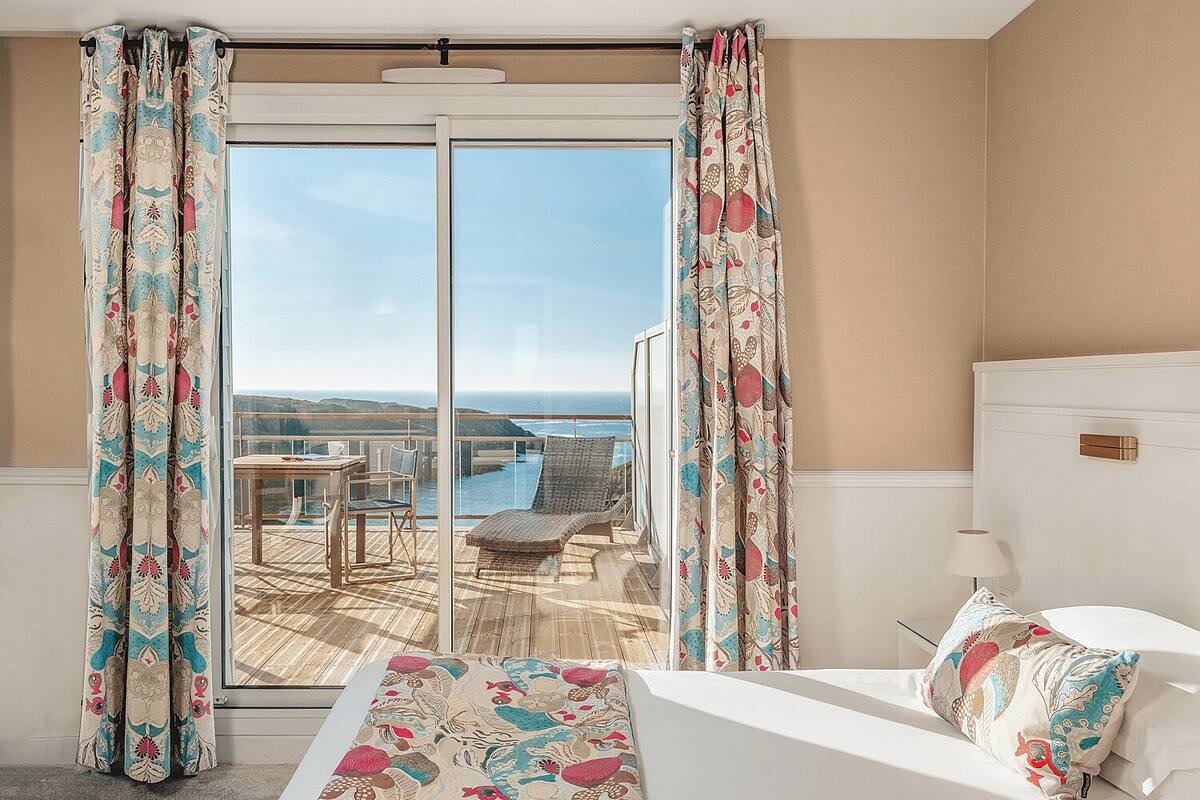 Quarto de hotel com varanda, cama com roupa de cama estampada e cortinas com motivos da natureza.
