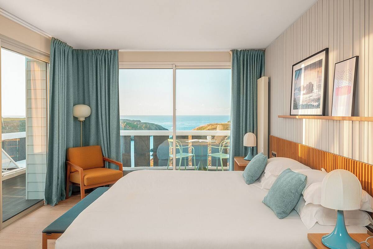 Quarto de hotel com cama grande, vista para o mar, varanda com cadeiras e cabeceira de madeira.