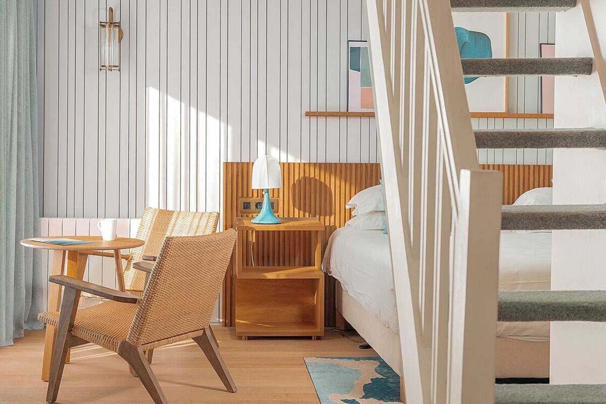 Quarto moderno com piso de madeira, uma cama, mesa de cabeceira, abajur e uma cadeira. Escada à direita.