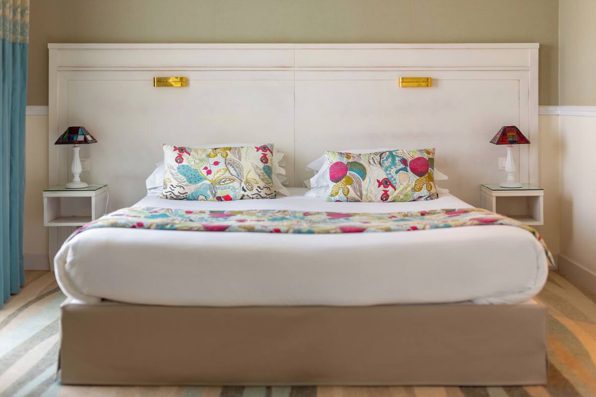 Cama bem arrumada com roupa de cama floral, duas mesas de cabeceira com abajures e uma cabeceira lisa.