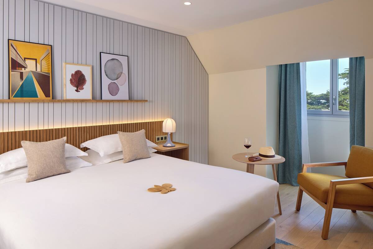 Quarto de hotel com uma cama grande, uma mesa pequena com abajur, uma cadeira e uma janela com cortinas.