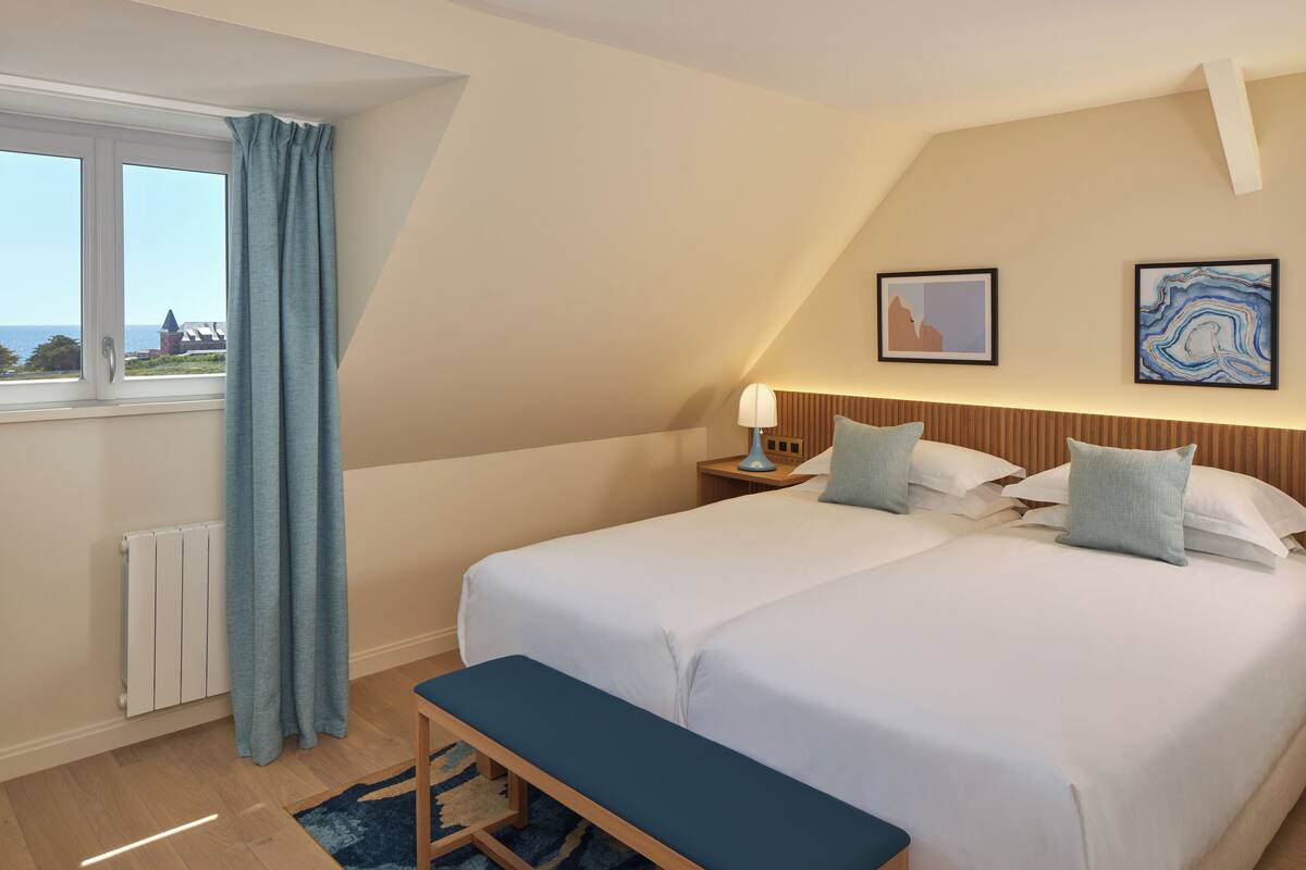 Quarto de hotel com uma cama grande, um banco, uma mesa de cabeceira com abajur e duas obras de arte emolduradas na parede.