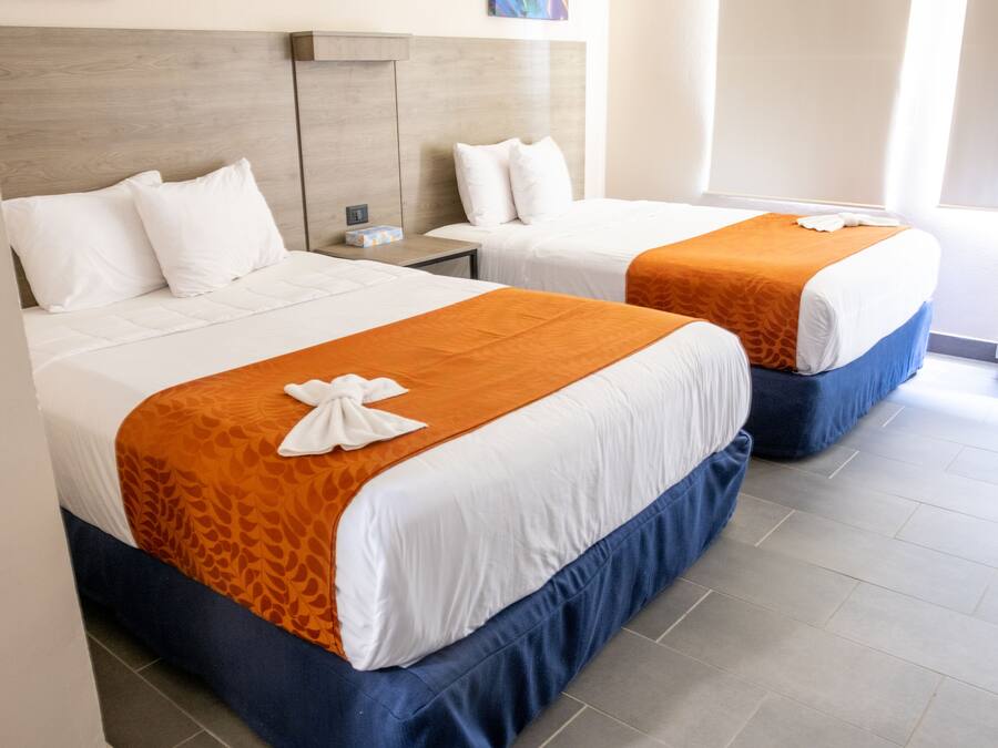 Quarto de hotel com duas camas, cada uma com roupa de cama branca e detalhes laranja, cabeceira de madeira, mesa de cabeceira e janela com persianas.