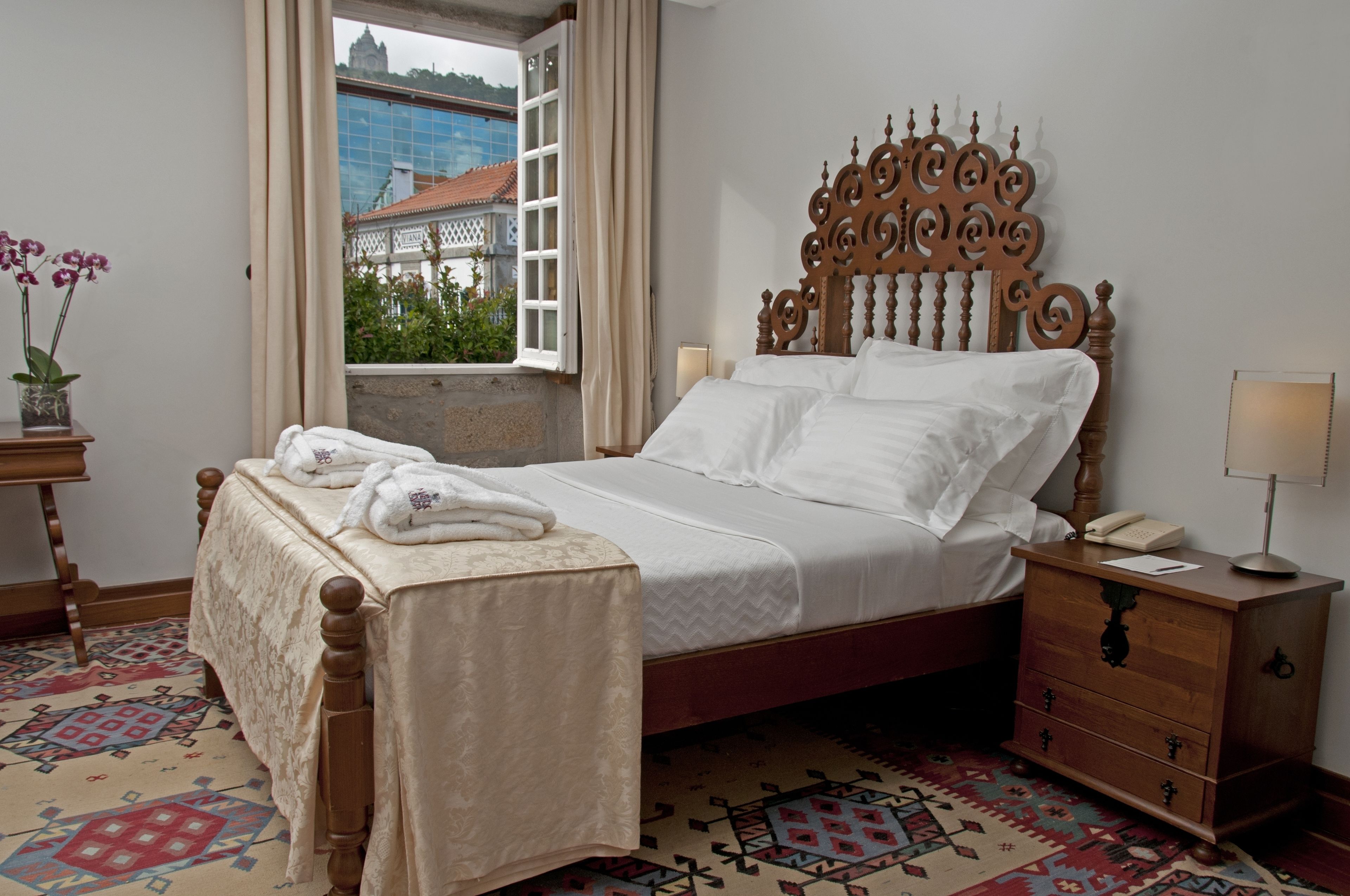 Uma cama bem arrumada com lençóis brancos, uma cabeceira de madeira, uma mesa de cabeceira com abajur e um tapete estampado.