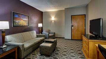 Quarto de hotel com sofá, poltronas, televisão, escrivaninha e carpete estampado.