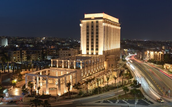 Hotel com uma torre imponente, área ajardinada com palmeiras e uma rua movimentada com rastros de luz.