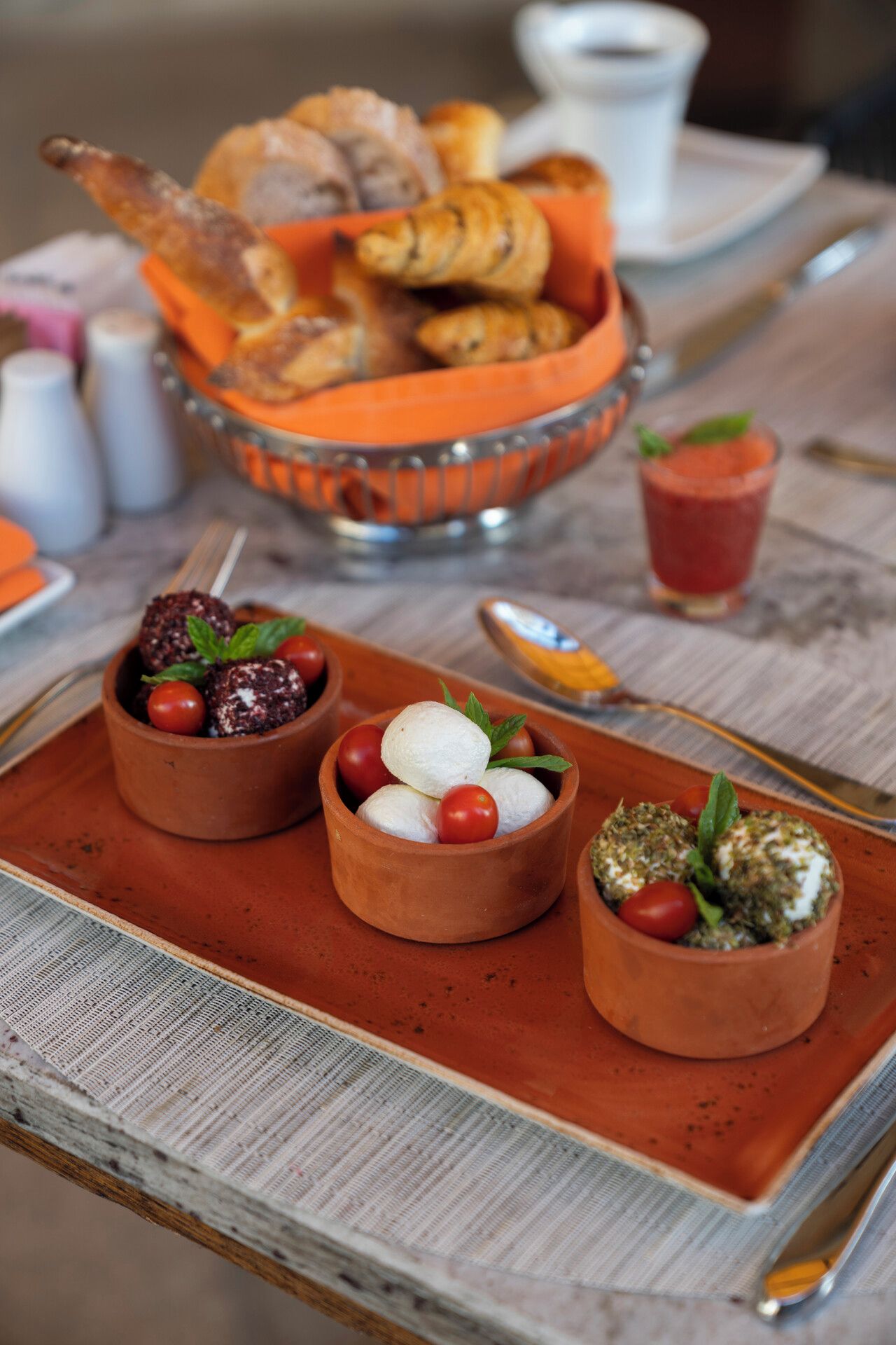 Uma mesa com diversos alimentos, incluindo uma cesta de pão, recipientes com diferentes pratos e uma chávena de bebida vermelha.