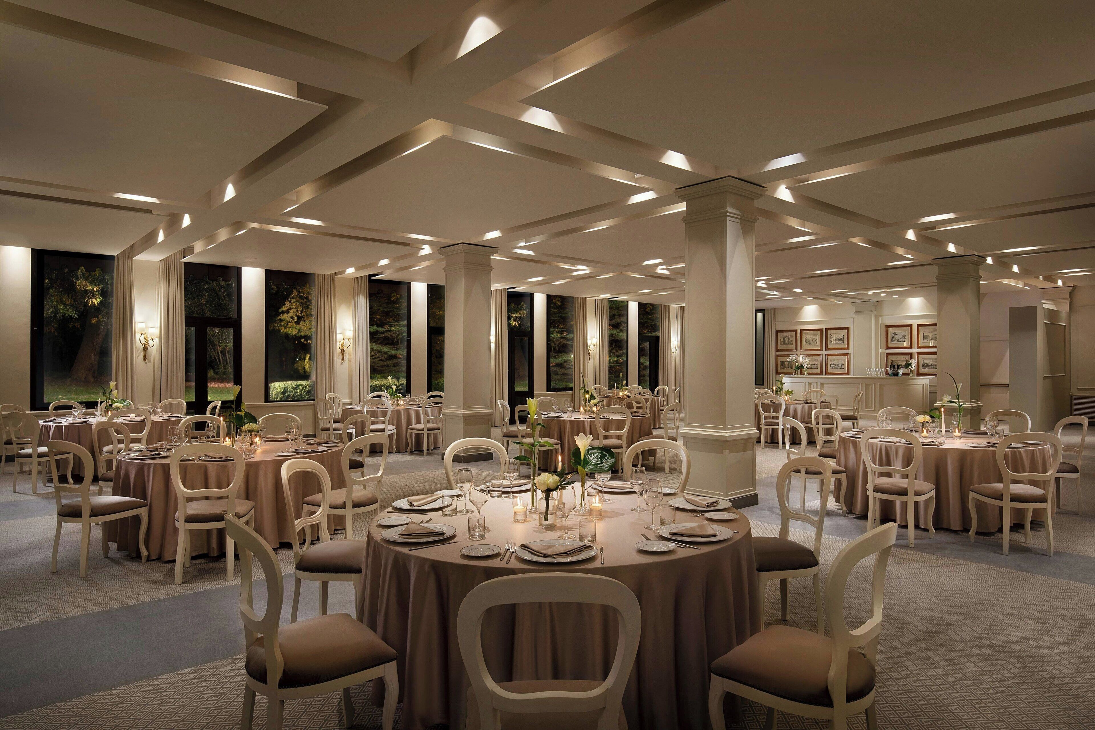 Uma ampla sala de banquetes com várias mesas redondas, cada uma com cadeiras e louças postas.