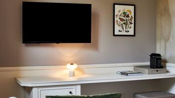 Quarto moderno com televisão de tela plana na parede, mesa de cabeceira com abajur, ilustração botânica emoldurada e máquina de café.