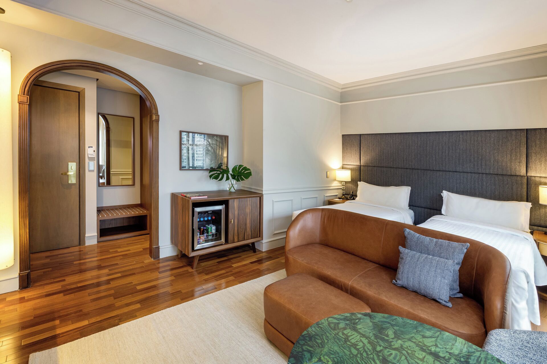 Quarto de hotel com cama, sofá, pequena geladeira, espelho e porta que dá para outro quarto.