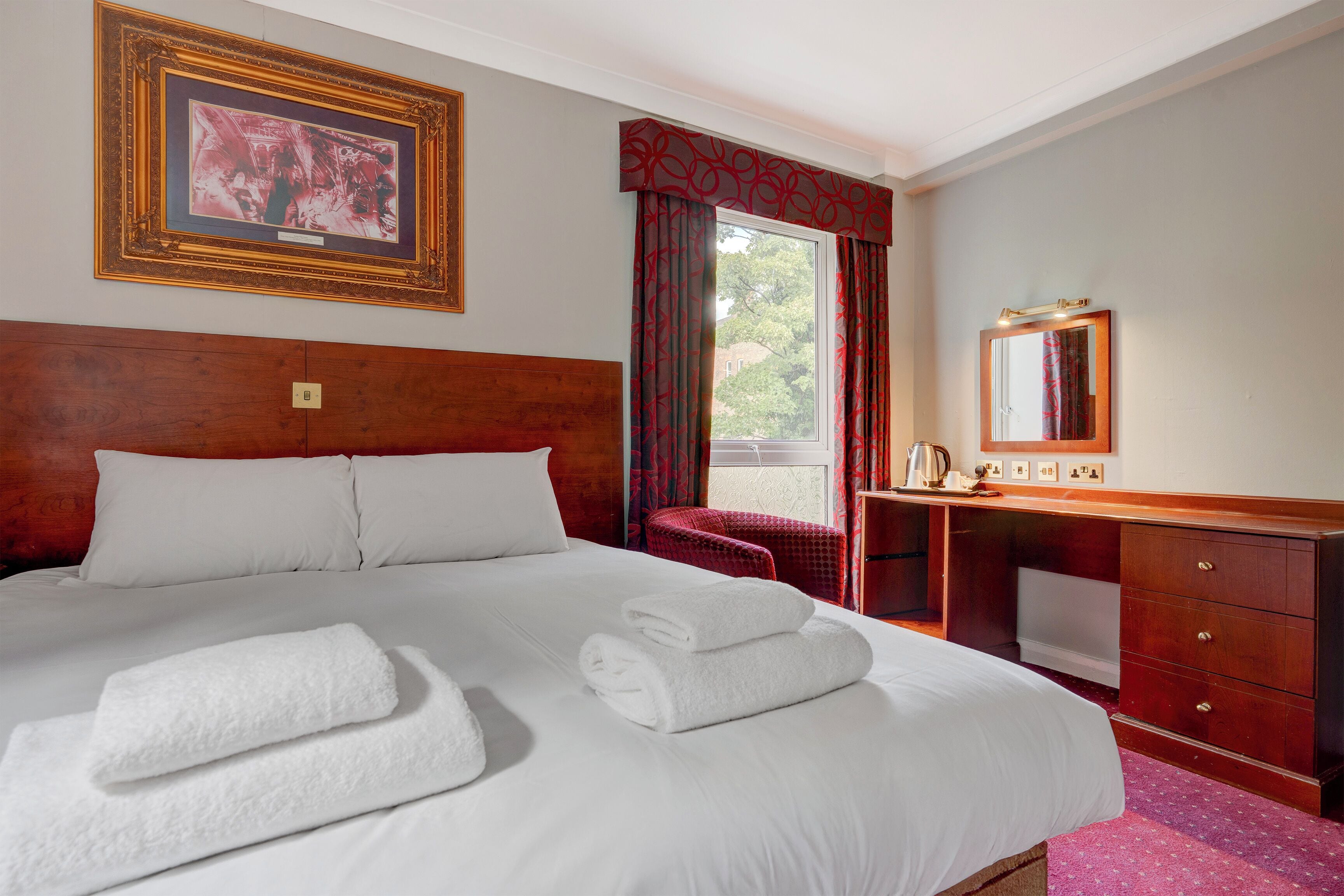 Quarto de hotel com uma cama grande, cabeceira de madeira, escrivaninha com espelho e janela com cortinas.