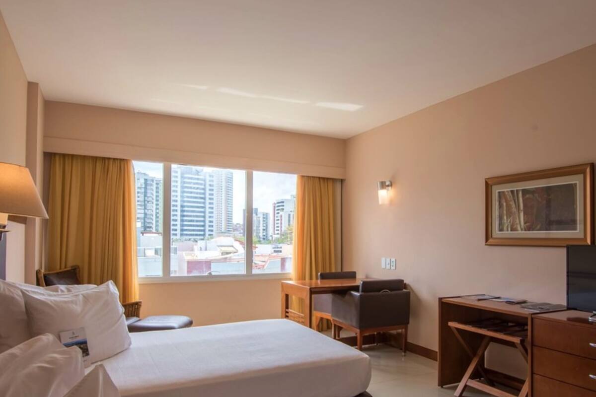 Quarto de hotel com cama, escrivaninha, cadeira, televisão e janela com vista para a cidade.
