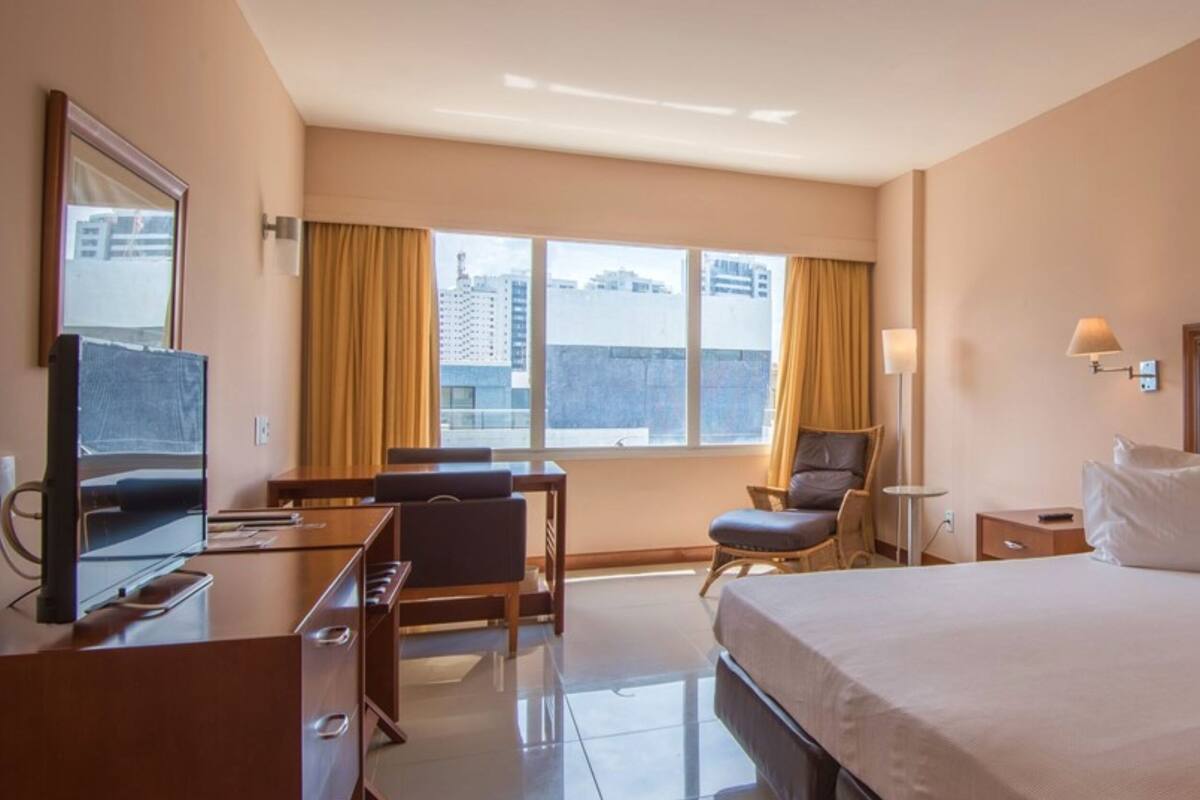 Quarto de hotel com uma cama, uma escrivaninha com computador, uma cadeira, televisão e janela com cortinas.