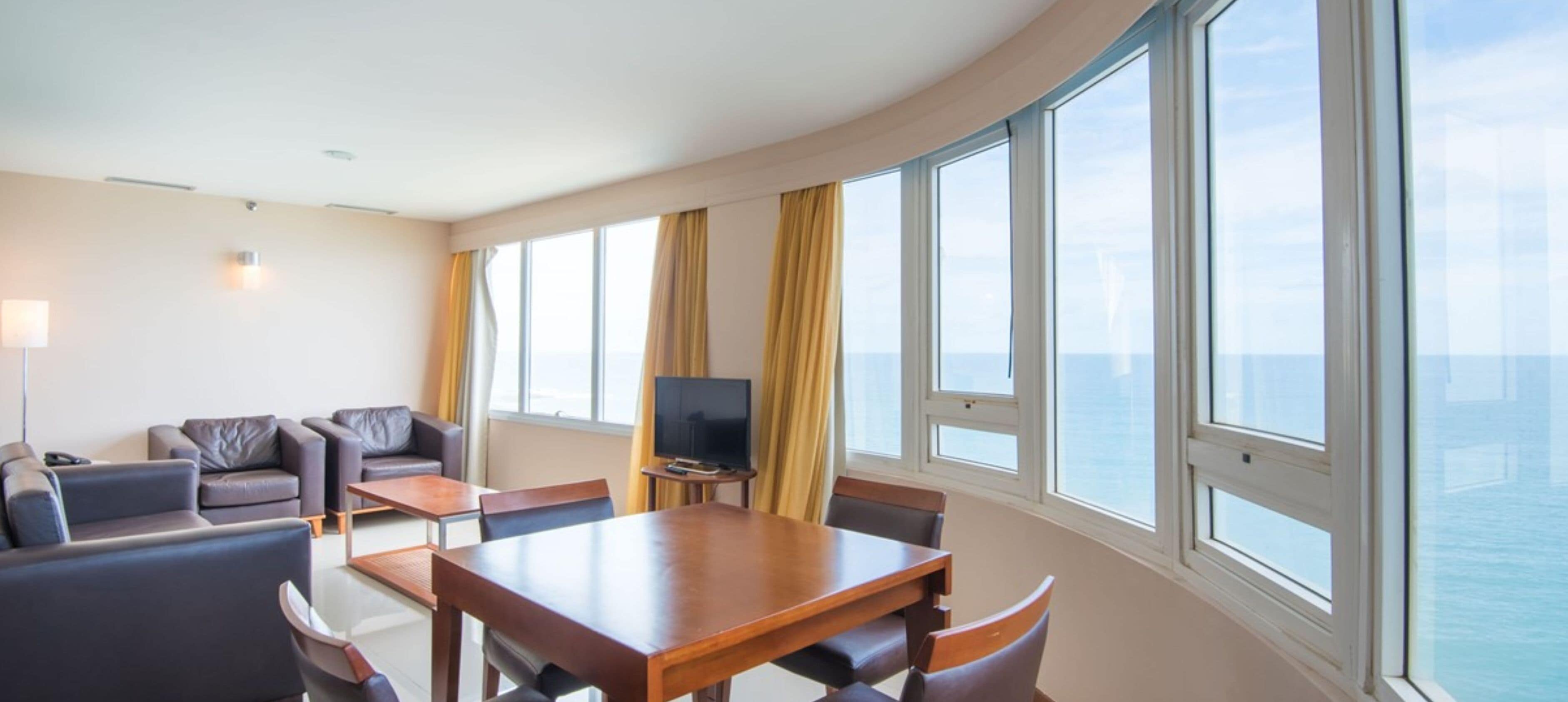 Quarto de hotel moderno com uma janela grande, área de estar com sofá e cadeiras, mesa de madeira e televisão de tela plana.