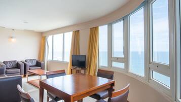 Quarto de hotel moderno com uma janela grande, área de estar com sofá e cadeiras, mesa de madeira e televisão de tela plana.
