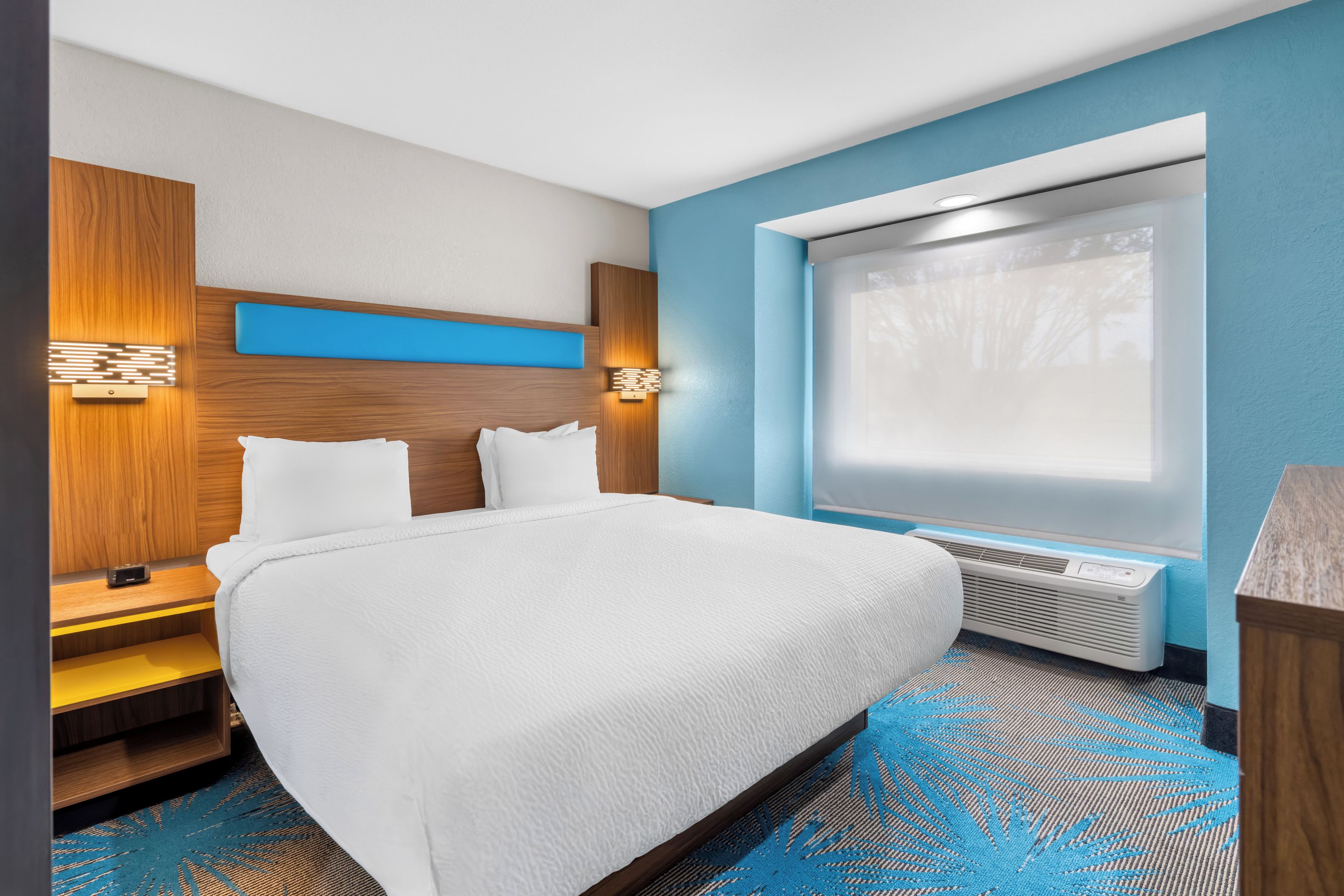Quarto de hotel com uma cama grande, uma mesinha de cabeceira, uma luminária na parede, janela com vista e ar condicionado integrado.