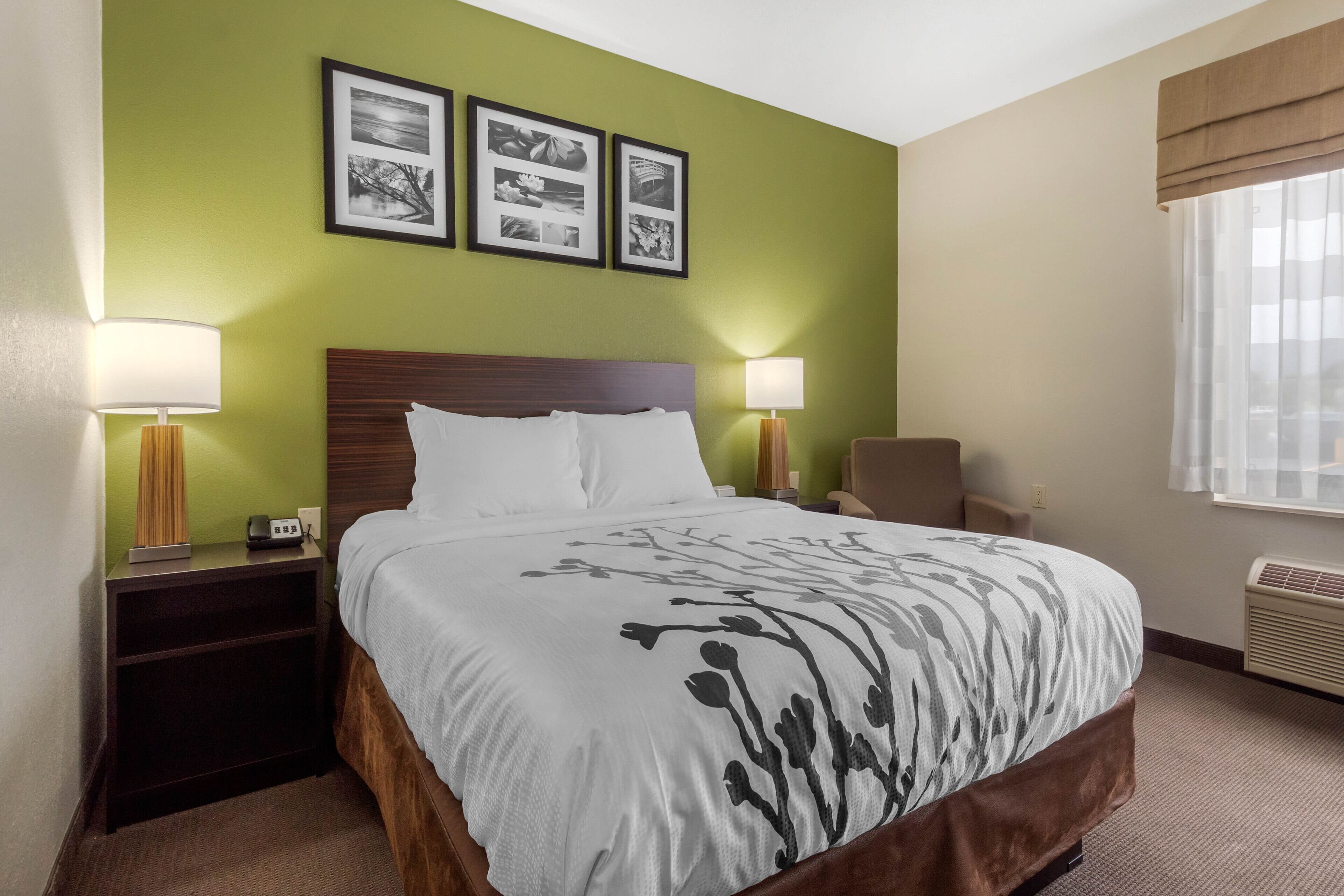 Quarto de hotel com cama grande, duas mesas de cabeceira com luminárias, uma cadeira e quadros nas paredes.