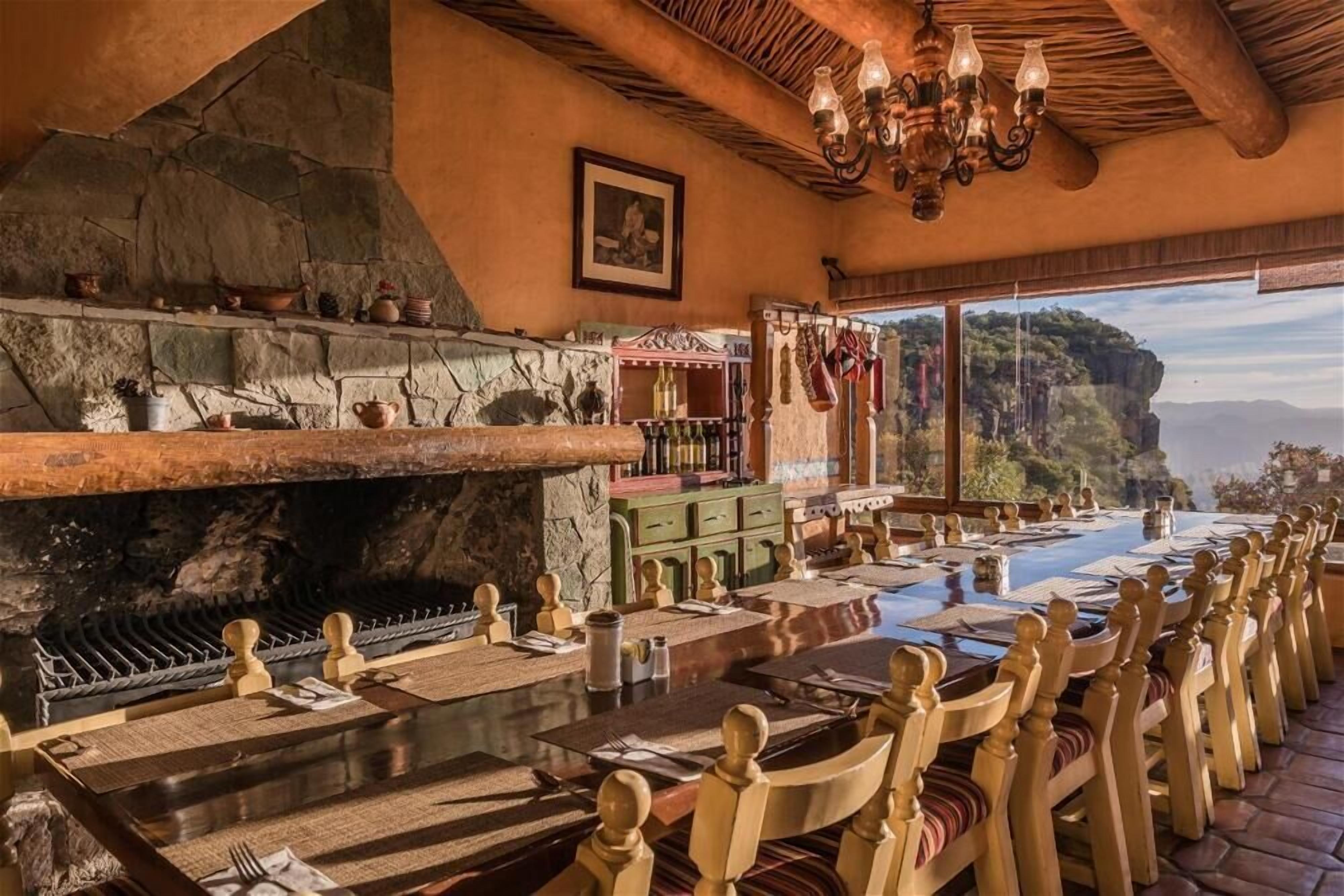 Uma sala de jantar com lareira de pedra, cadeiras de madeira e uma mesa posta para refeição.