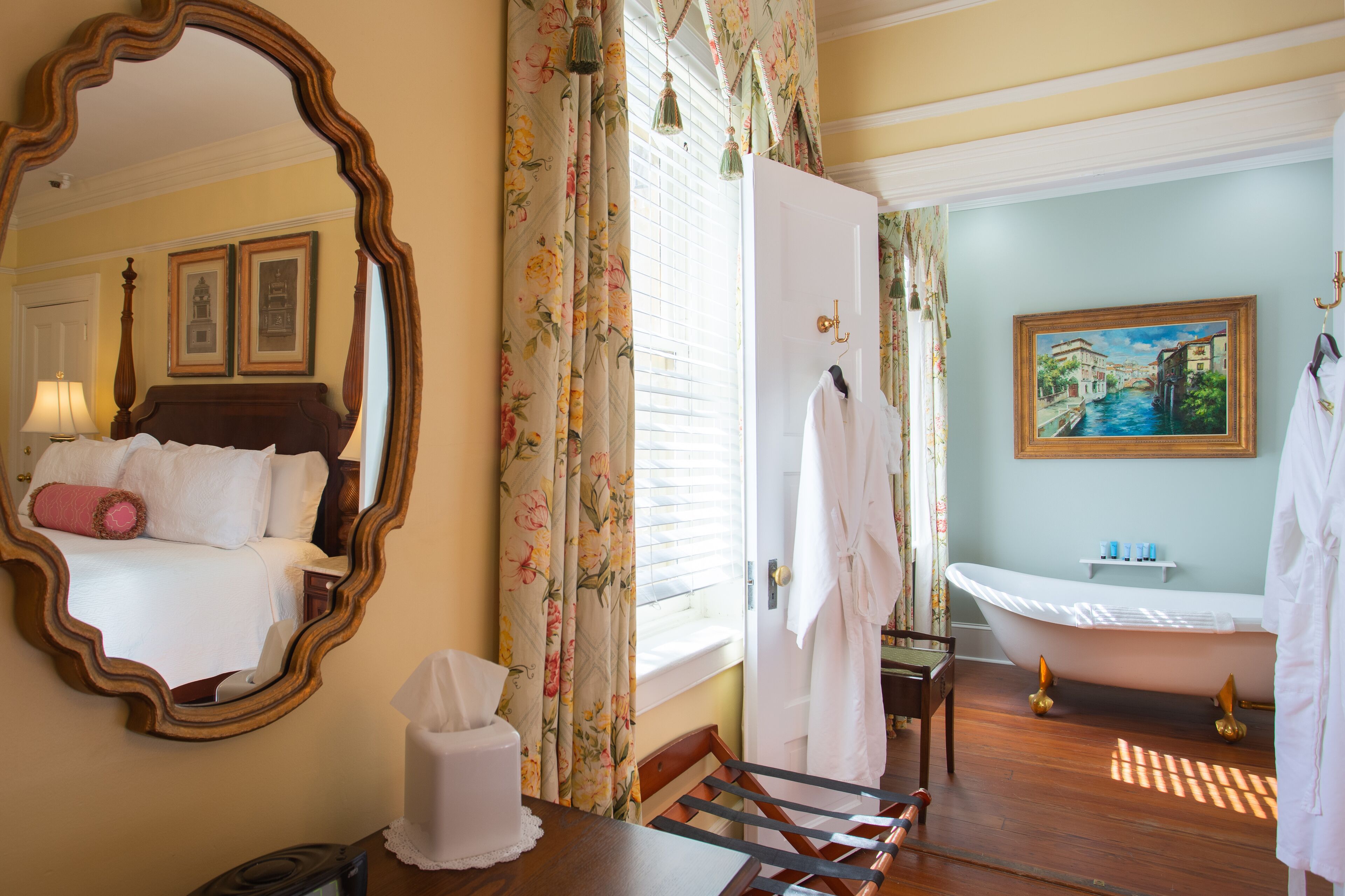 Quarto de hotel com cama, um espelho grande, banheiro com banheira e um quadro na parede.