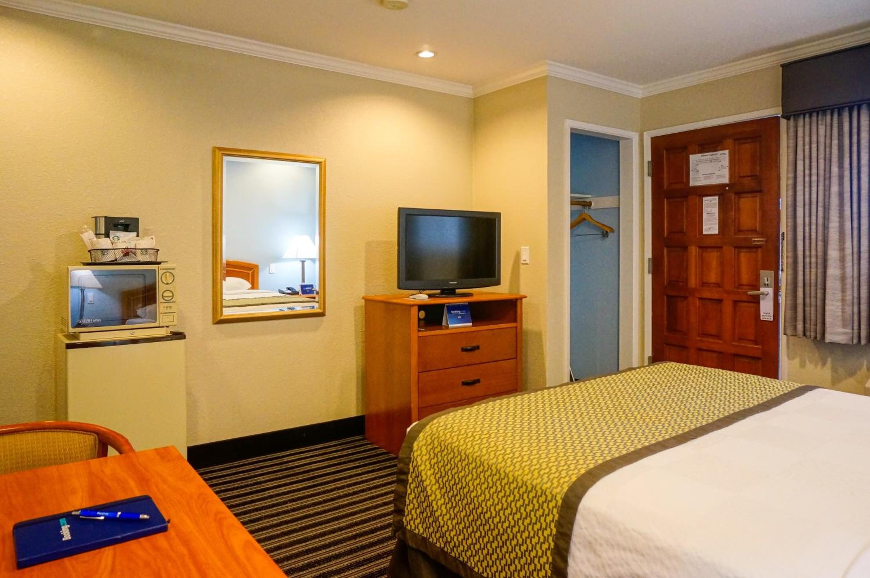 Quarto de hotel com uma cama, uma televisão sobre um suporte de madeira, uma escrivaninha com uma cadeira e um espelho na parede.