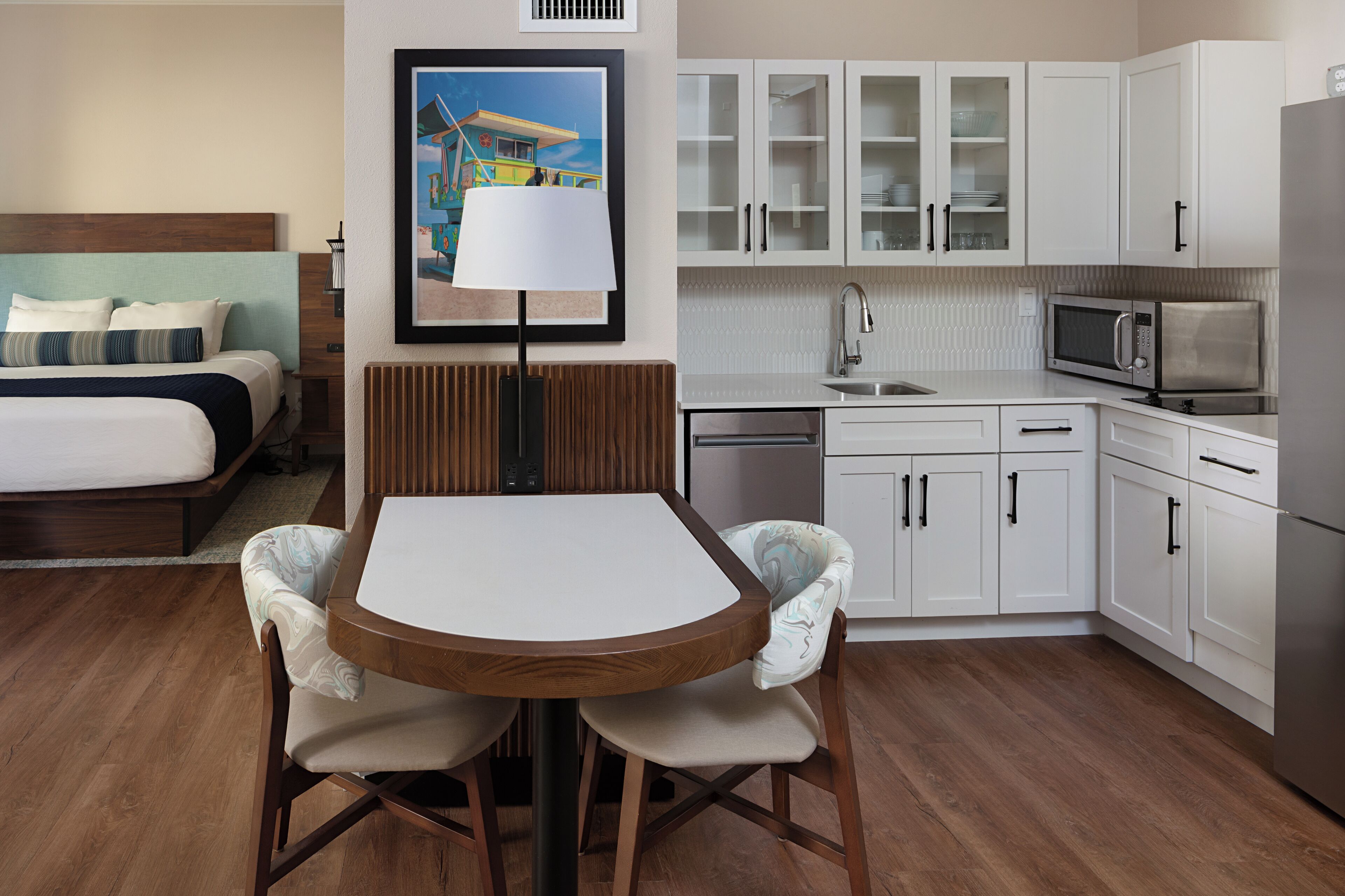 Quarto de hotel moderno com piso de madeira, cama com cabeceira, mesa de jantar com cadeiras, cozinha com armários e eletrodomésticos brancos, e uma moldura com um quadro na parede.