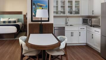Quarto de hotel moderno com piso de madeira, cama com cabeceira, mesa de jantar com cadeiras, cozinha com armários e eletrodomésticos brancos, e uma moldura com um quadro na parede.