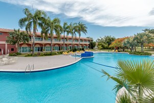 Piscina de resort com águas cristalinas e azuis, rodeada por palmeiras e um edifício com varandas.