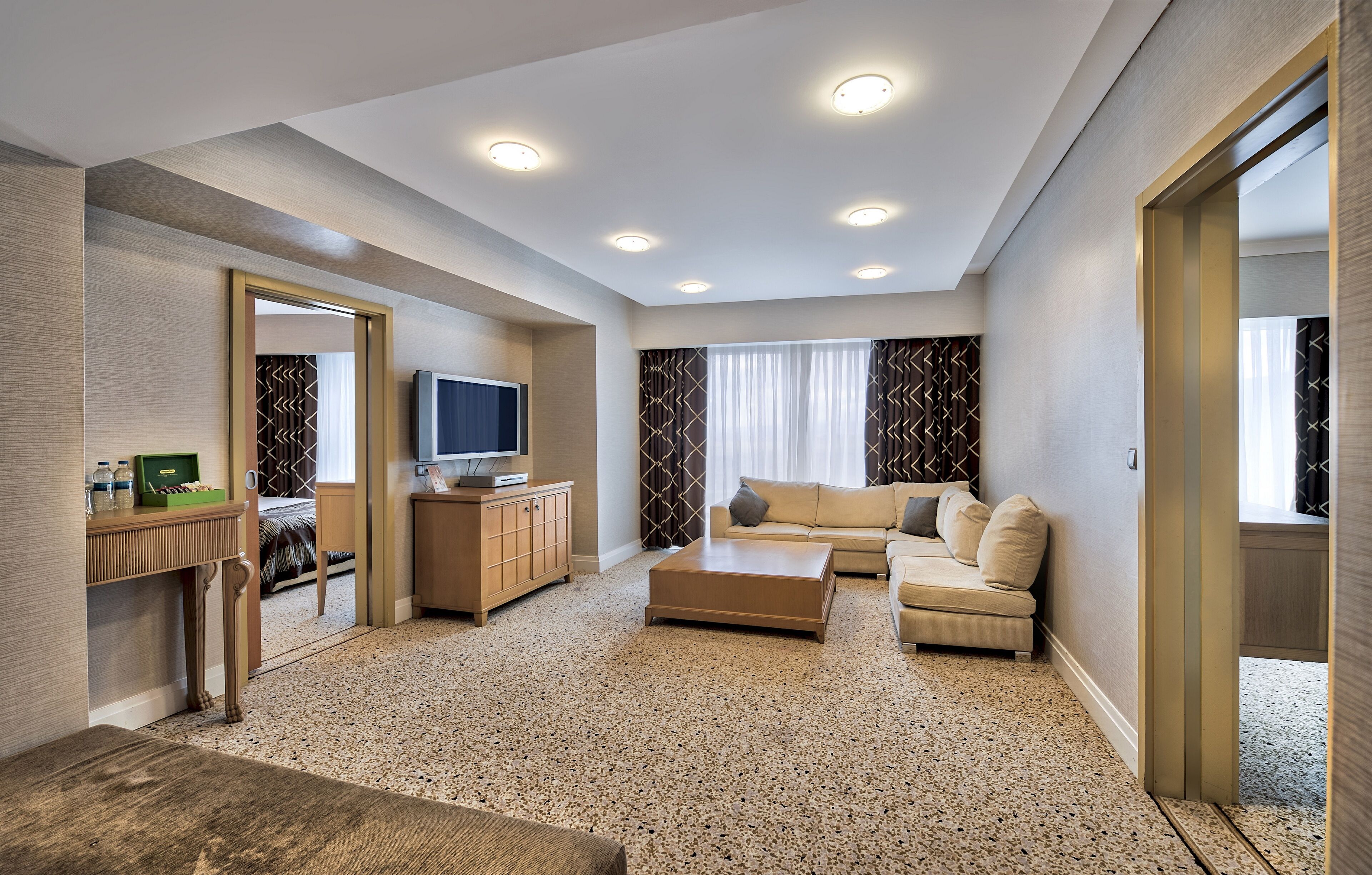 Quarto de hotel moderno com área de estar, incluindo sofá, mesa de centro e televisão. Há uma escrivaninha de madeira com espelho e armário. O quarto possui piso coberto por carpete e parede com padrão.