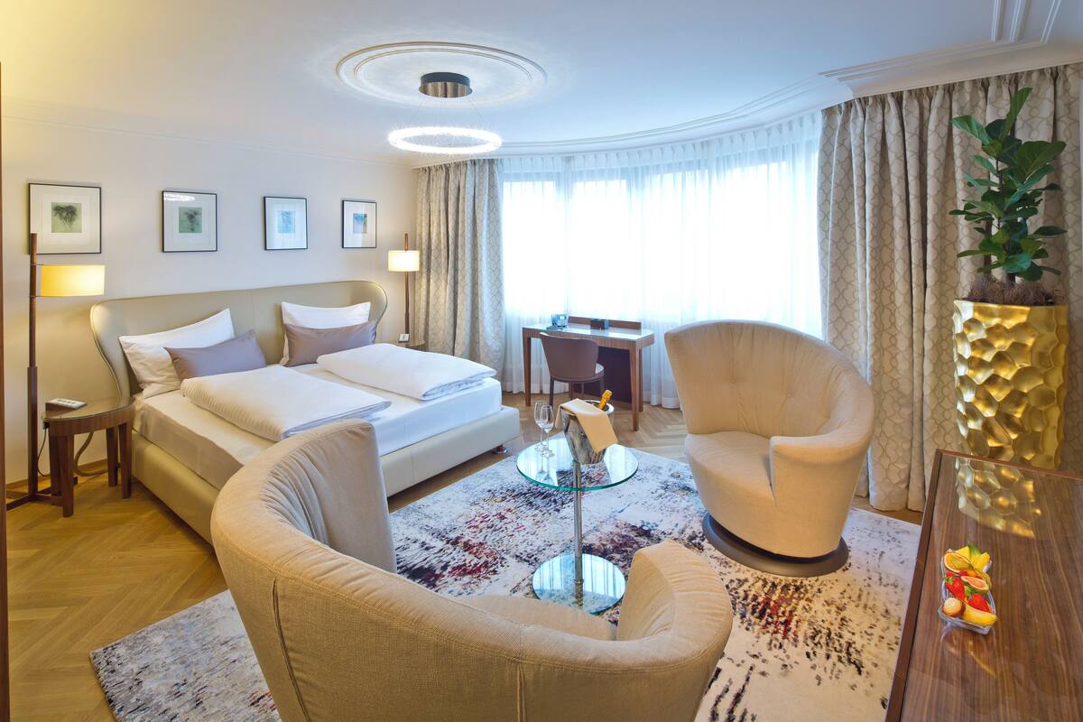 Quarto de hotel com cama, área de estar com poltrona e mesa, escrivaninha e uma janela grande com cortinas.