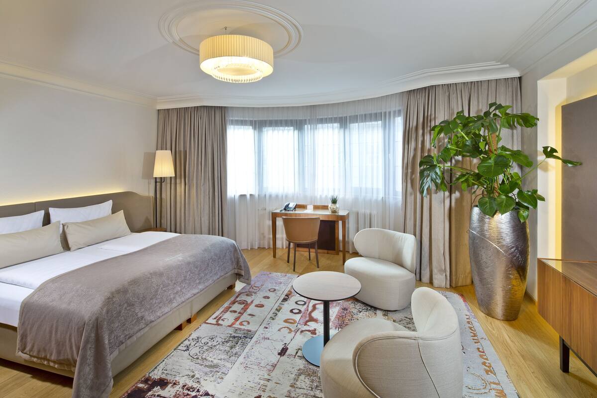 Quarto de hotel com cama, escrivaninha, poltrona, sofá, abajur e uma planta grande.