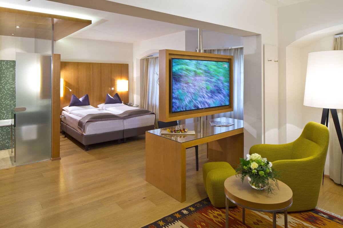 Quarto de hotel com cama, televisão, escrivaninha, cadeira e uma pequena mesa com um vaso de flores.