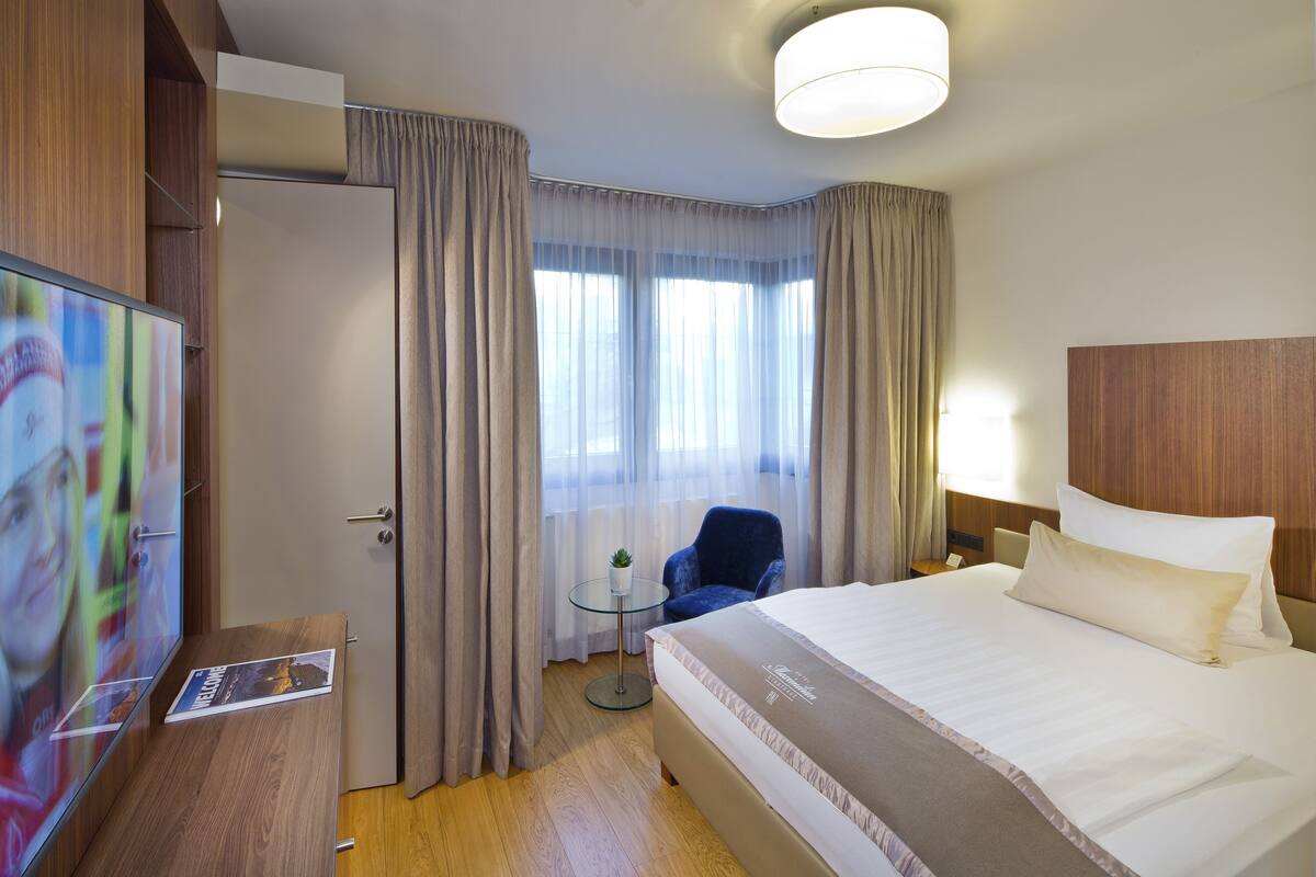 Quarto de hotel com cama, escrivaninha, cadeira, mesa pequena e televisão.