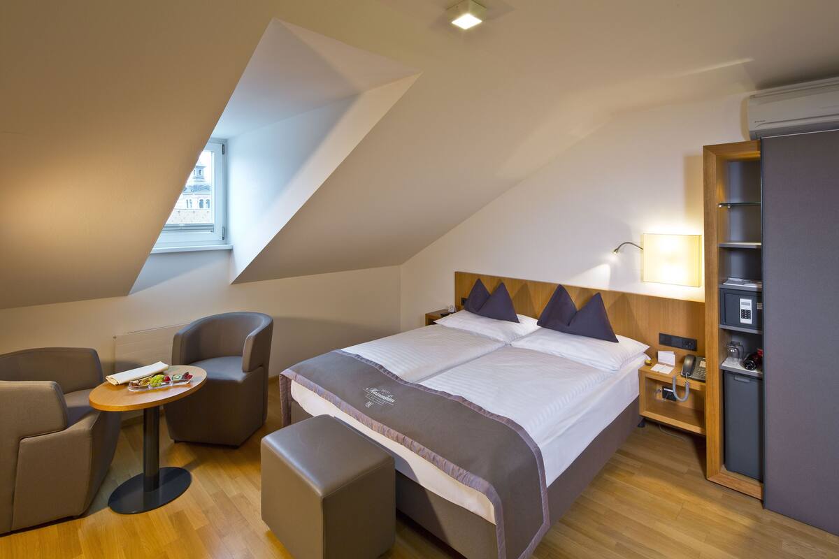 Quarto de hotel moderno com uma cama grande, uma escrivaninha, uma cadeira e uma pequena mesa com uma bandeja de frutas.