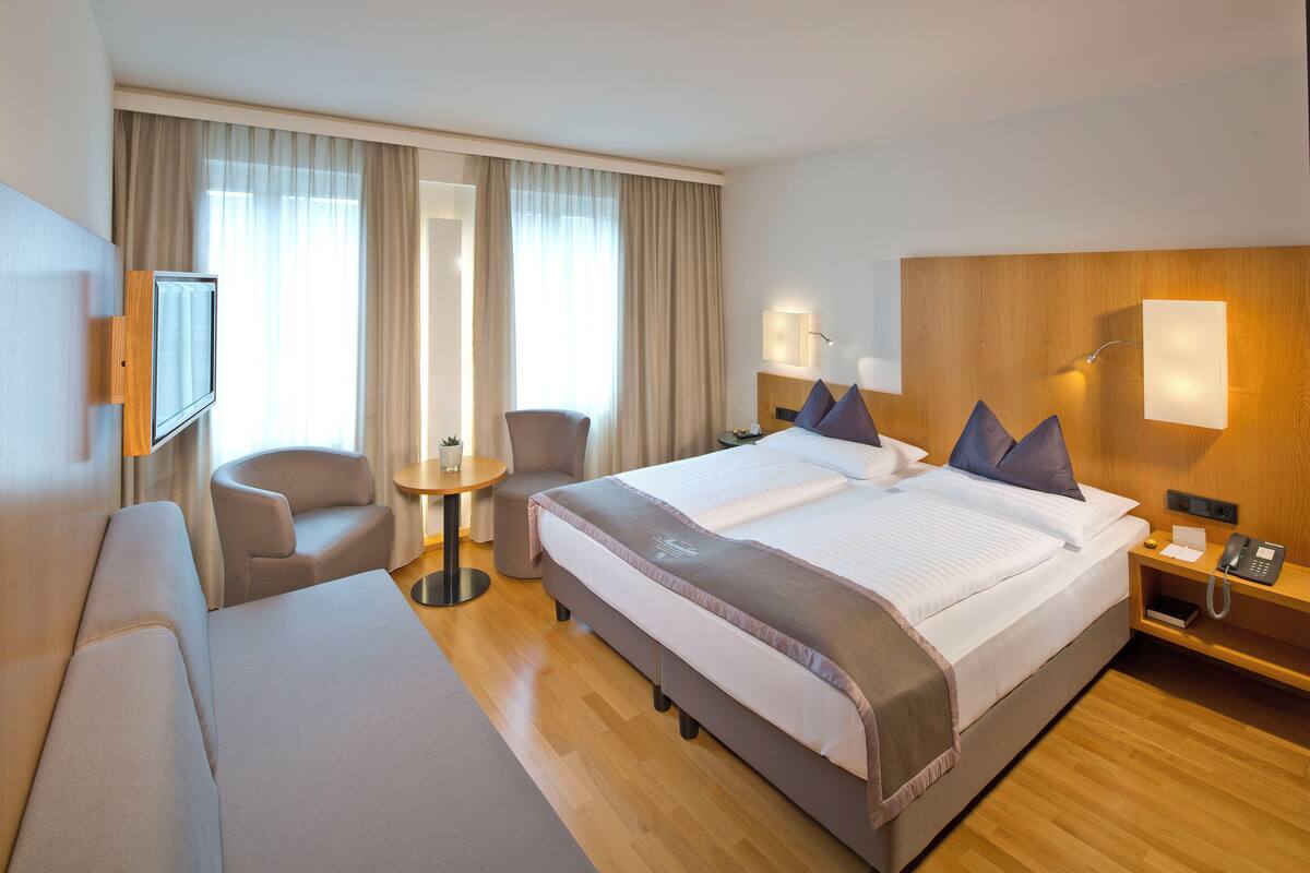 Quarto de hotel com uma cama grande, um sofá, uma poltrona, uma mesa pequena e uma mesinha de cabeceira com luminária e telefone.