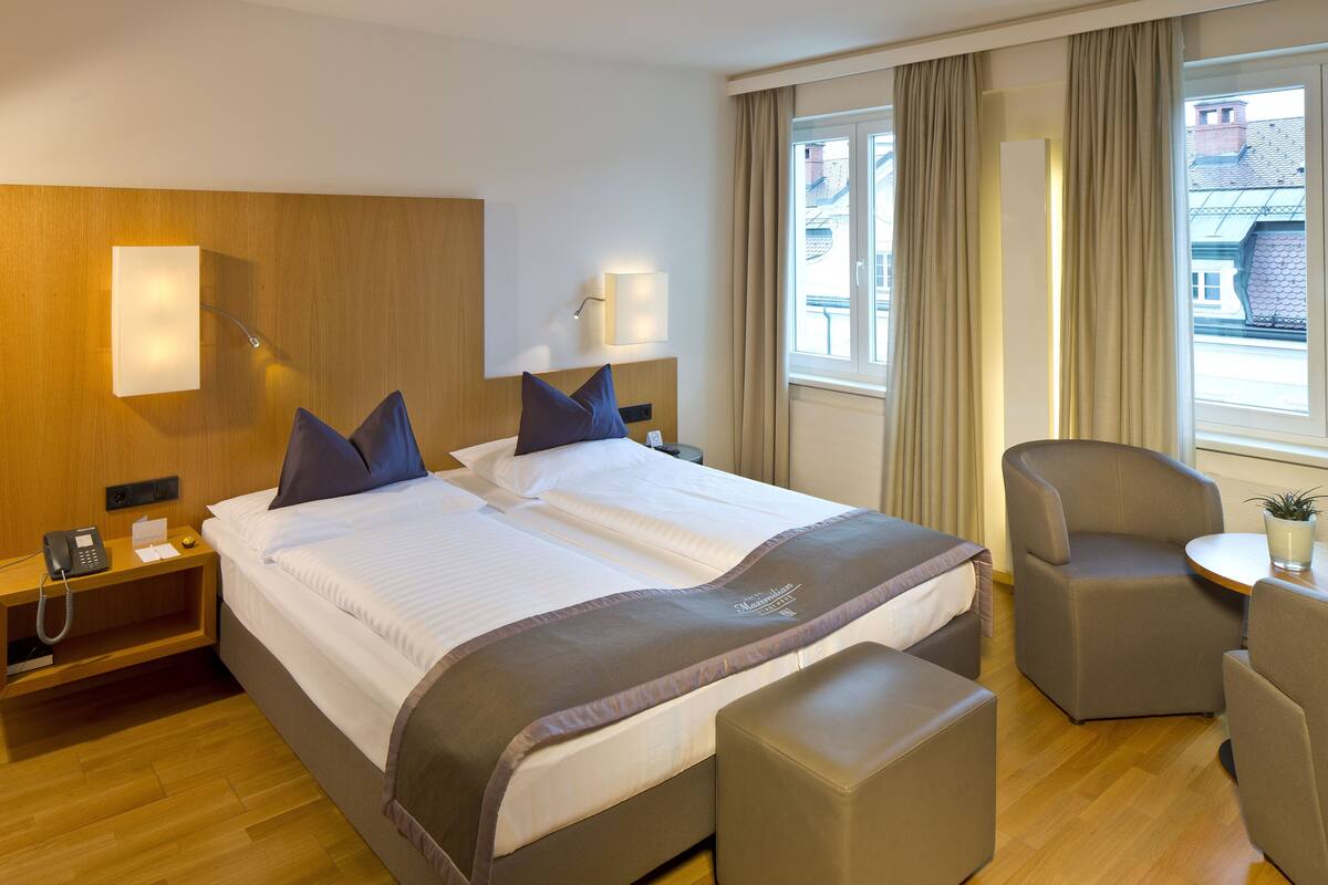 Quarto de hotel com uma cama grande, duas mesas de cabeceira, uma escrivaninha, uma cadeira e uma janela com cortinas.