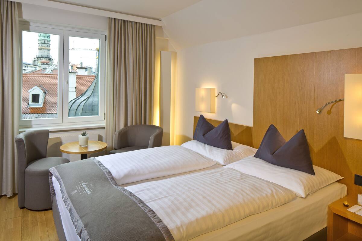 Quarto de hotel com duas camas, uma mesinha, uma cadeira e uma janela com cortinas.