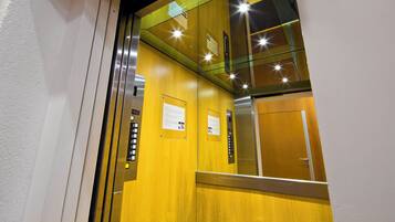 Interior de elevador com paredes de madeira, guarda-mão metálico e painel de comandos com botões.