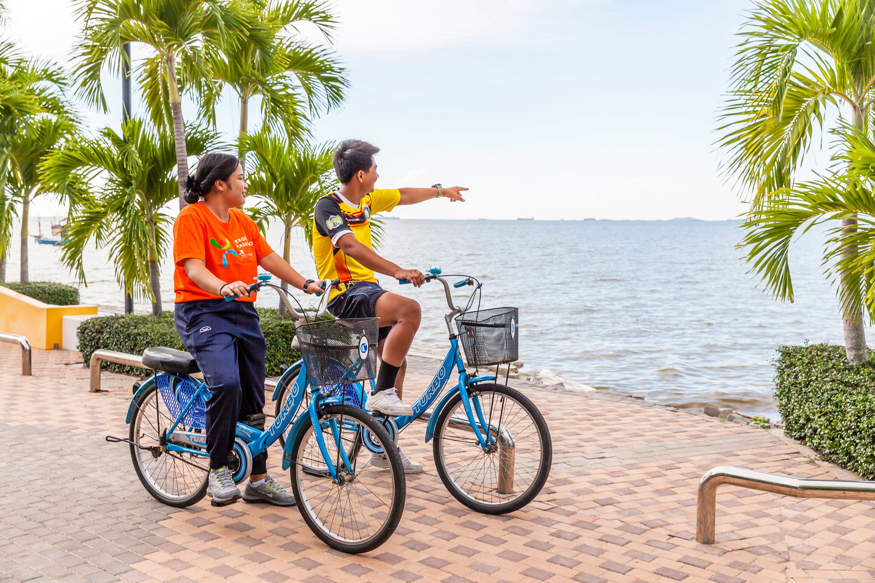 Duas pessoas em uma bicicleta azul com cestas, perto de um corpo d'água e palmeiras.