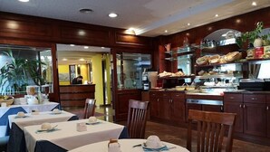 Restaurante com mesas redondas preparadas para refeições, área de cozinha com vitrines e um balcão de recepção.