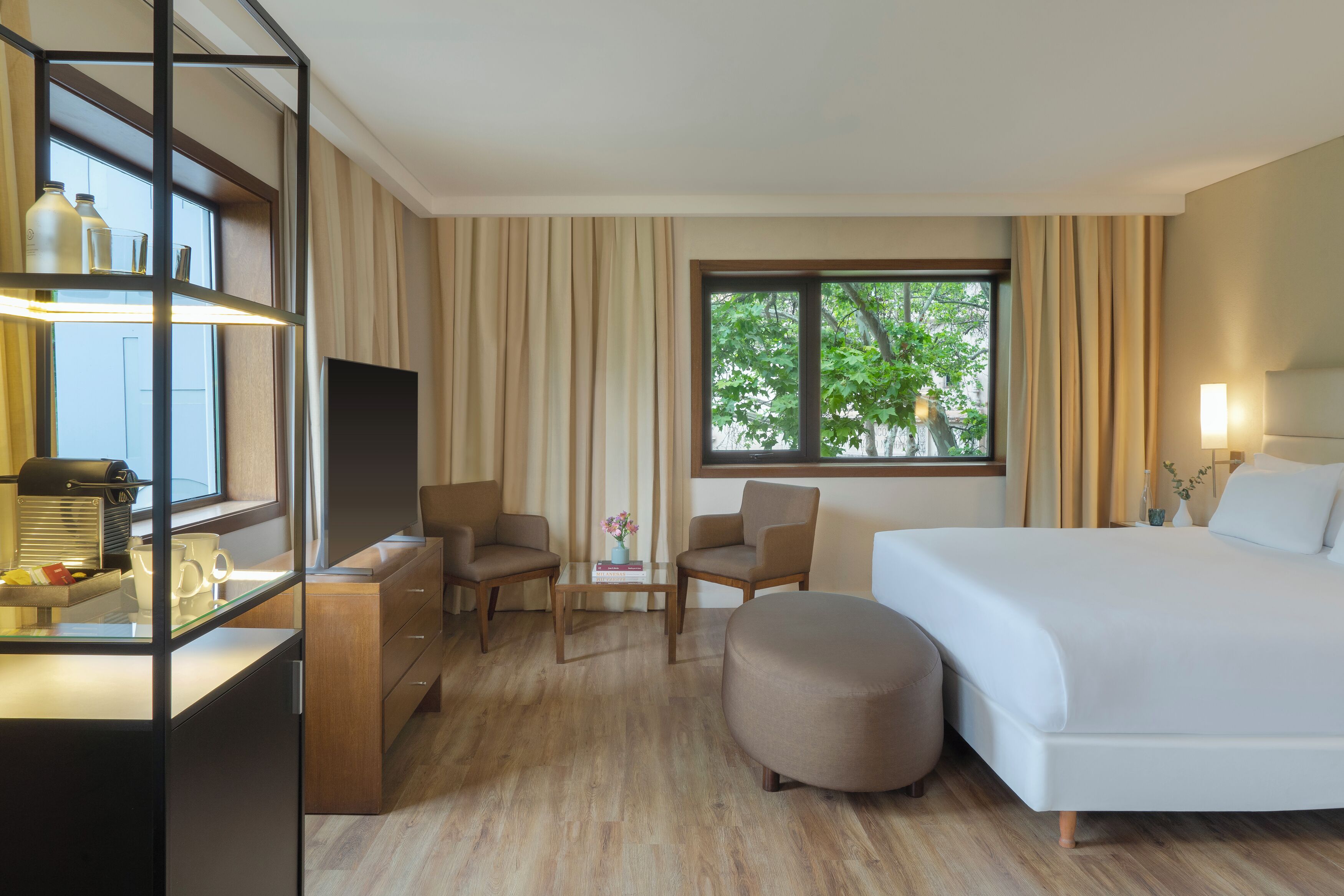 Quarto de hotel com uma cama grande, duas poltronas, uma mesa pequena, televisão e uma janela com cortinas.