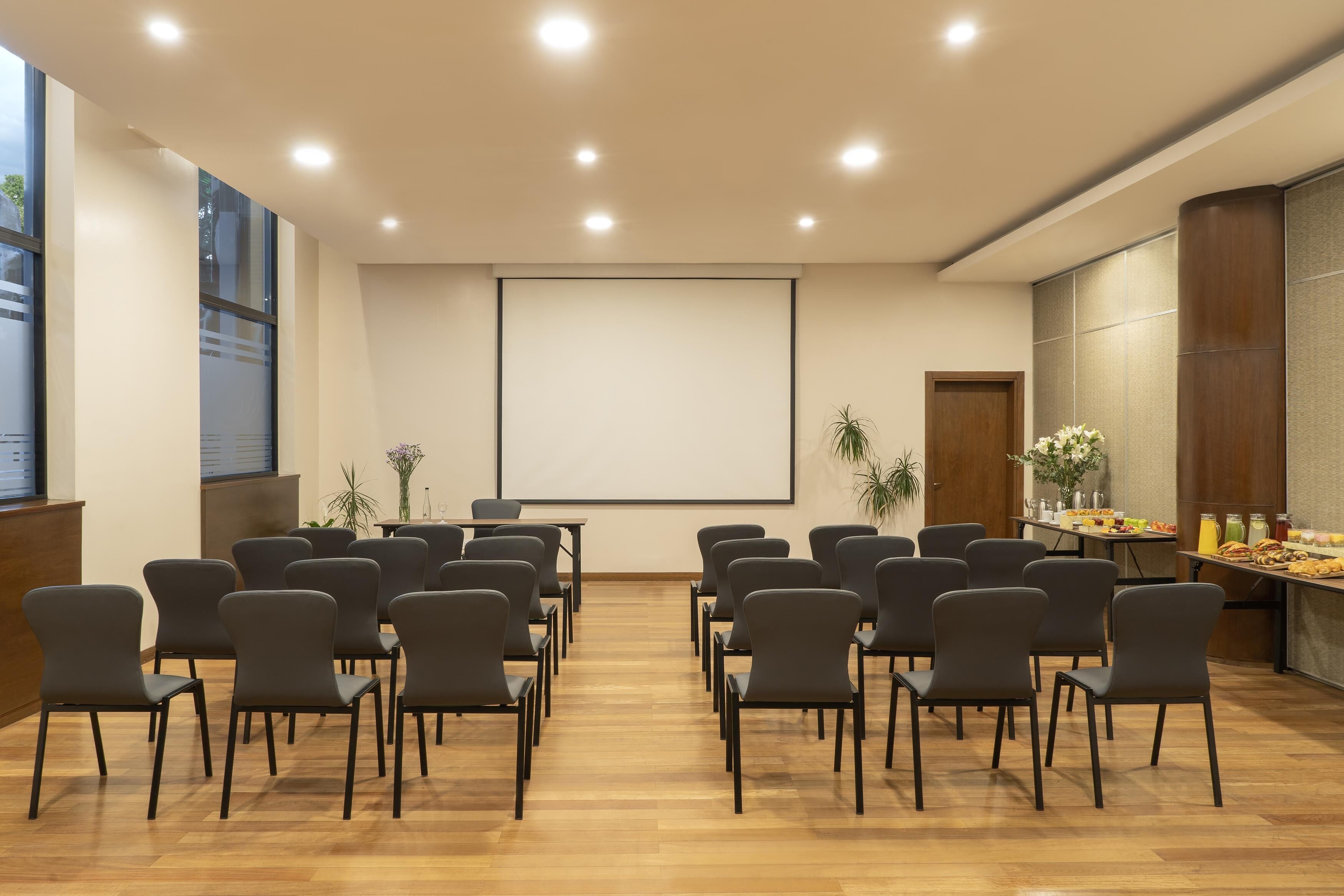 Uma sala de conferências com fileiras de cadeiras voltadas para uma tela de projeção, um balcão de recepção em madeira e uma mesa de buffet com comida e bebidas.