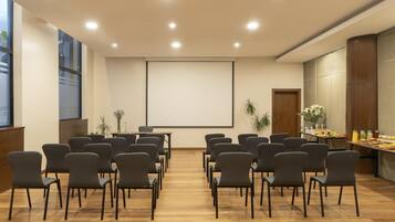 Uma sala de conferências com fileiras de cadeiras voltadas para uma tela de projeção, um balcão de recepção em madeira e uma mesa de buffet com comida e bebidas.