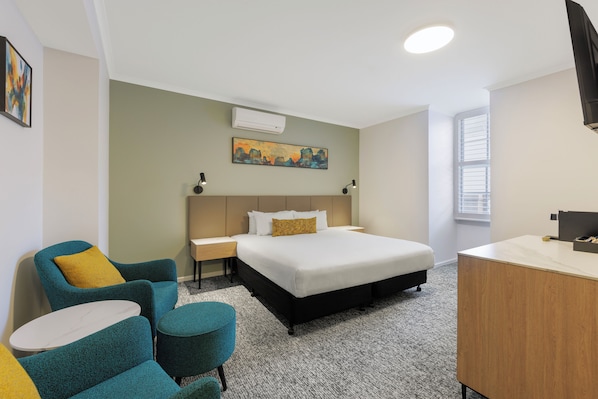 Quarto de hotel com cama, escrivaninha, cadeira, mesa pequena e um quadro na parede.