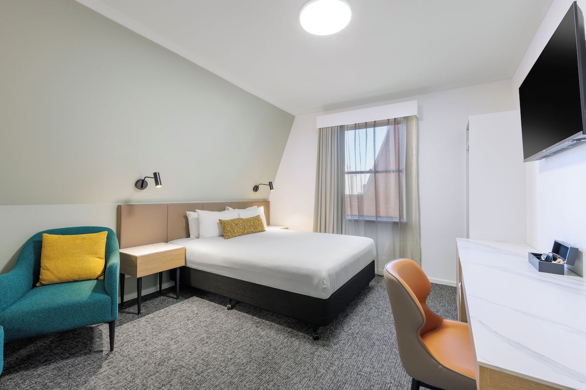 Quarto de hotel com cama, escrivaninha, cadeira e televisão.