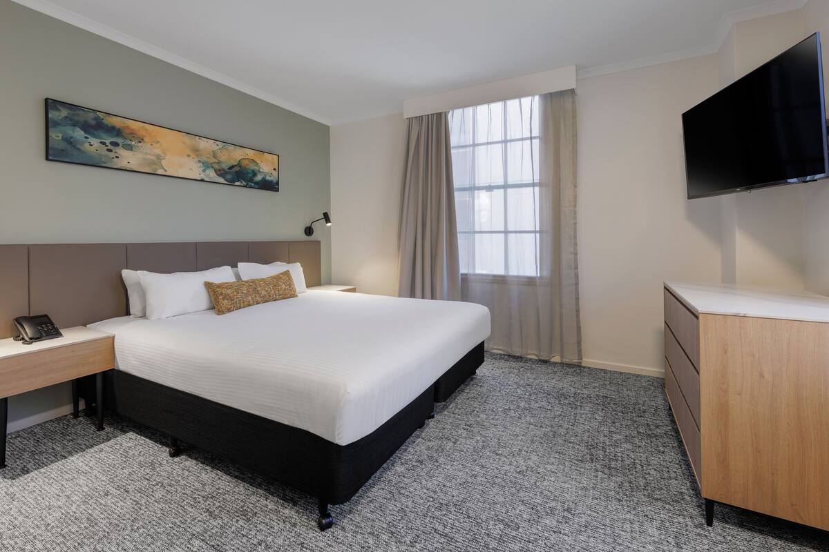 Quarto de hotel com uma cama grande, mesa de cabeceira com telefone, cômoda, televisão de tela plana e janela com cortinas.