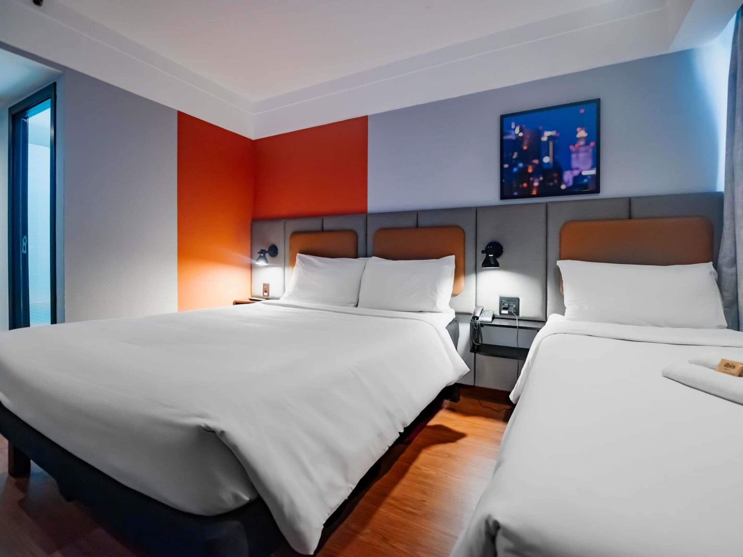 Quarto de hotel com duas camas, uma mesa de cabeceira com abajur, um quadro na parede e uma porta à esquerda.