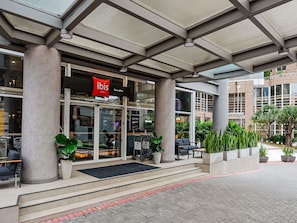 Entrada de hotel com galeria coberta, colunas imponentes e área de estar externa.