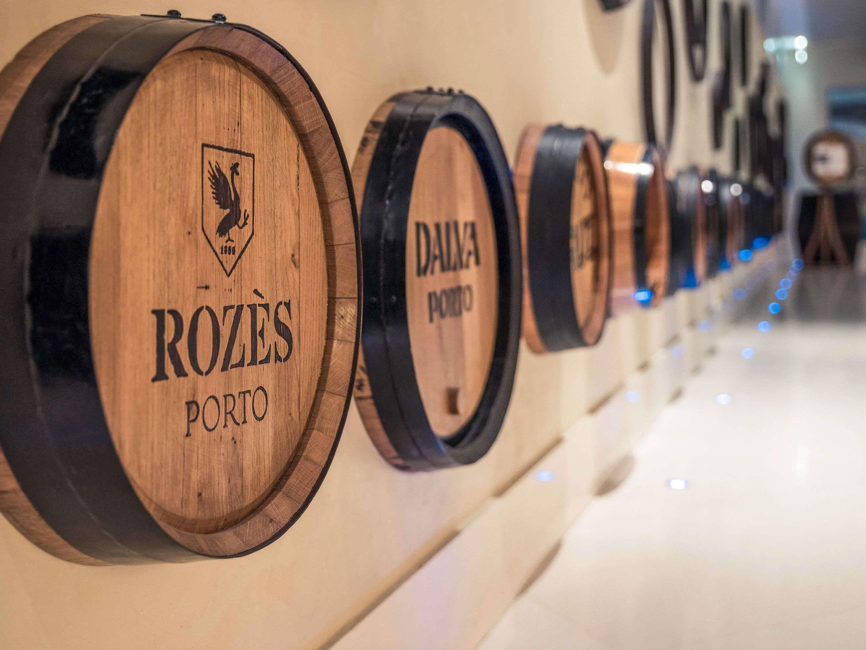 Fileira de barris de madeira com etiquetas, provavelmente para armazenamento de vinho, em ambiente interno bem iluminado.