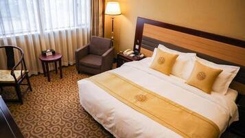 Quarto de hotel com uma cama grande, uma cadeira, uma mesinha de cabeceira, um abajur e uma pequena mesa.
